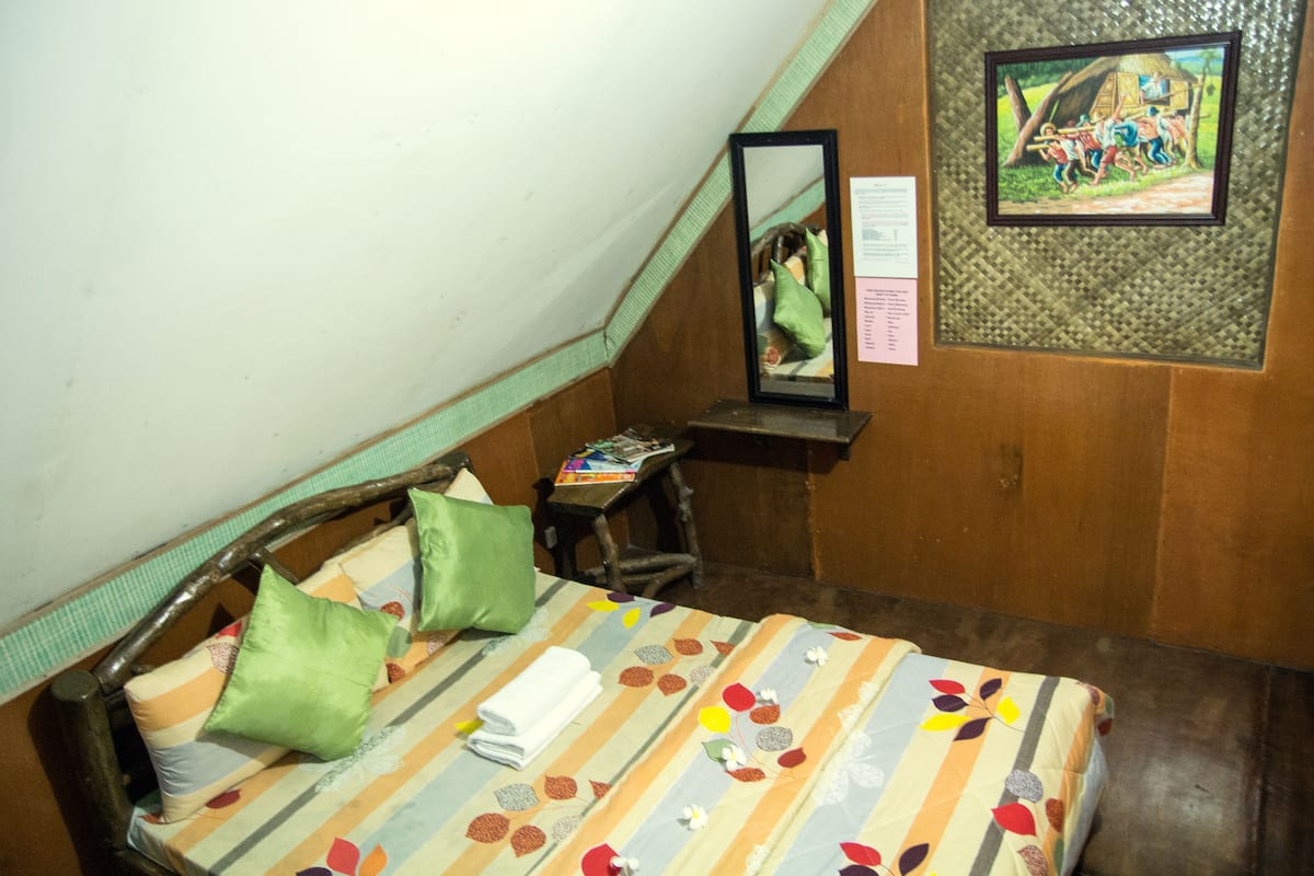 Quarto com cama, almofadas, uma mesinha de cabeceira com espelho e um quadro na parede.