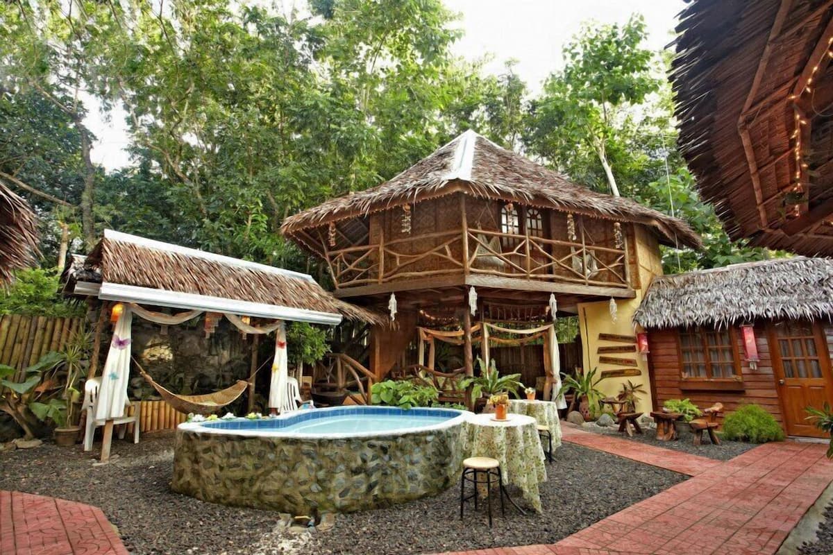 Ambiente rústico ao ar livre, com uma estrutura de telhado de palha, uma pequena piscina e um deck de madeira.