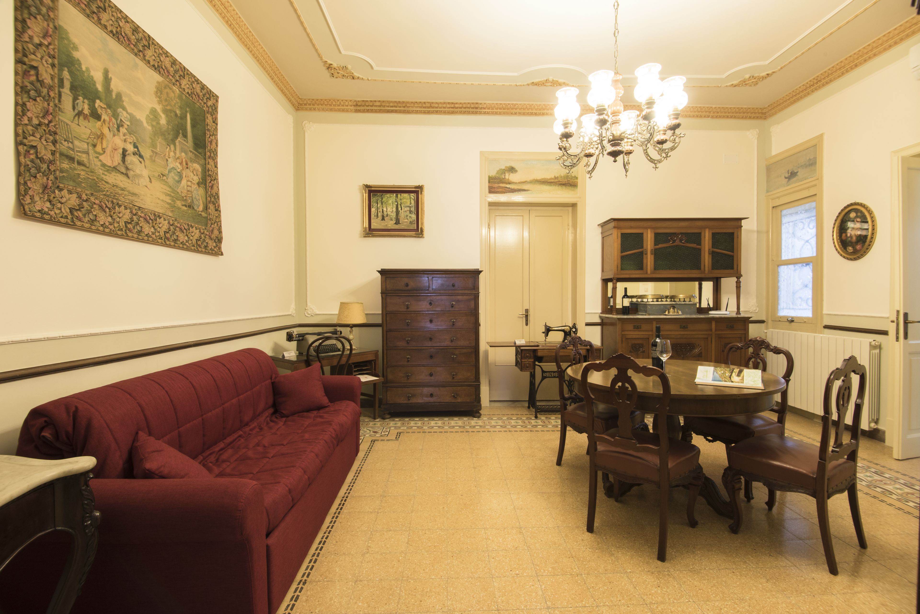 Uma sala de jantar com mesa e cadeiras de madeira, um sofá vermelho, um lustre e um quadro na parede.