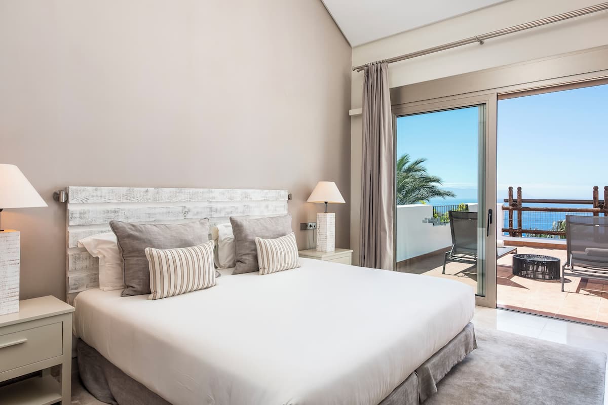 Quarto de hotel com uma cama grande, duas mesas de cabeceira com abajures, vista para o mar e uma varanda com área de estar ao ar livre.