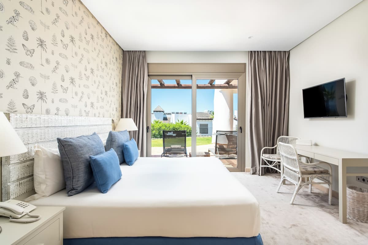 Quarto de hotel com uma cama grande, uma escrivaninha, uma cadeira, televisão e varanda com vista para edifícios e vegetação.