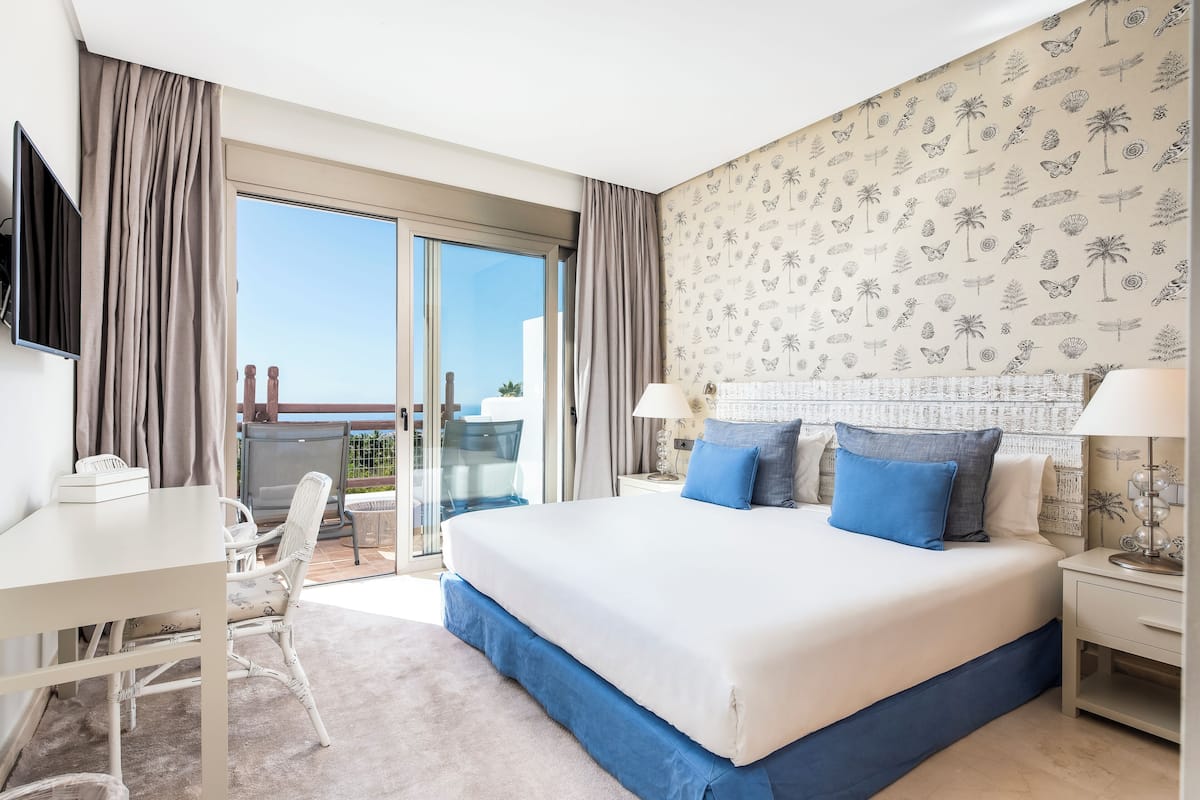 Quarto de hotel com cama grande, uma mesa, uma cadeira, televisão e varanda com porta de vidro.