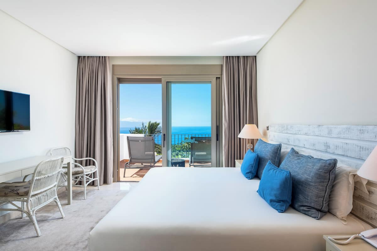 Quarto de hotel com uma cama grande, uma escrivaninha, uma cadeira e uma televisão. Possui uma varanda com vista para o oceano.