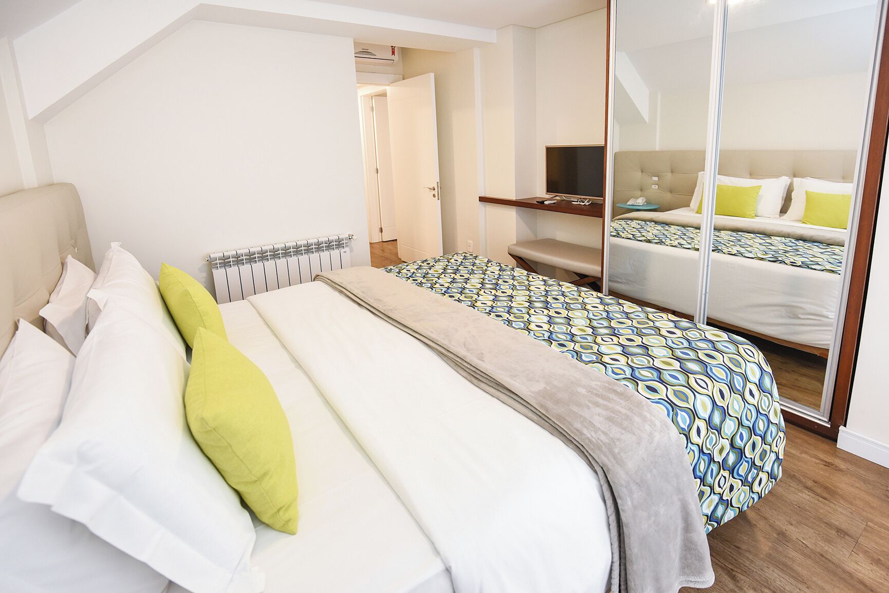 Quarto de hotel moderno com uma cama grande, mesa de cabeceira, espelho e guarda-roupa.