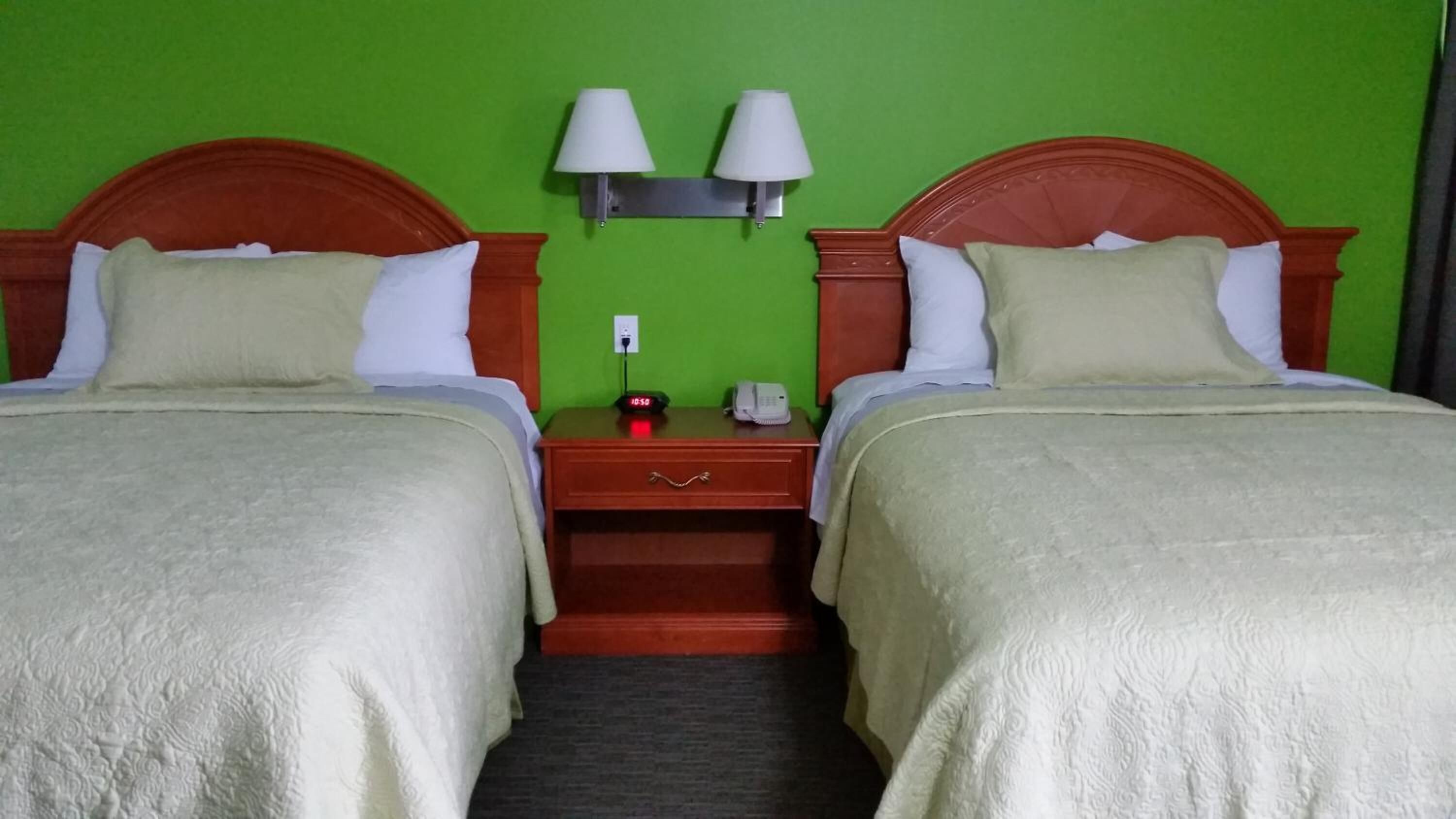 Quarto de hotel com duas camas, cada uma com cabeceira, mesa de cabeceira com relógio e telefone, e luminária fixada na parede.