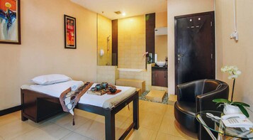 Quarto com cama, mesa de massagem, cadeira, pequena mesa com telefone e planta, e banheiro visível através de uma porta aberta.