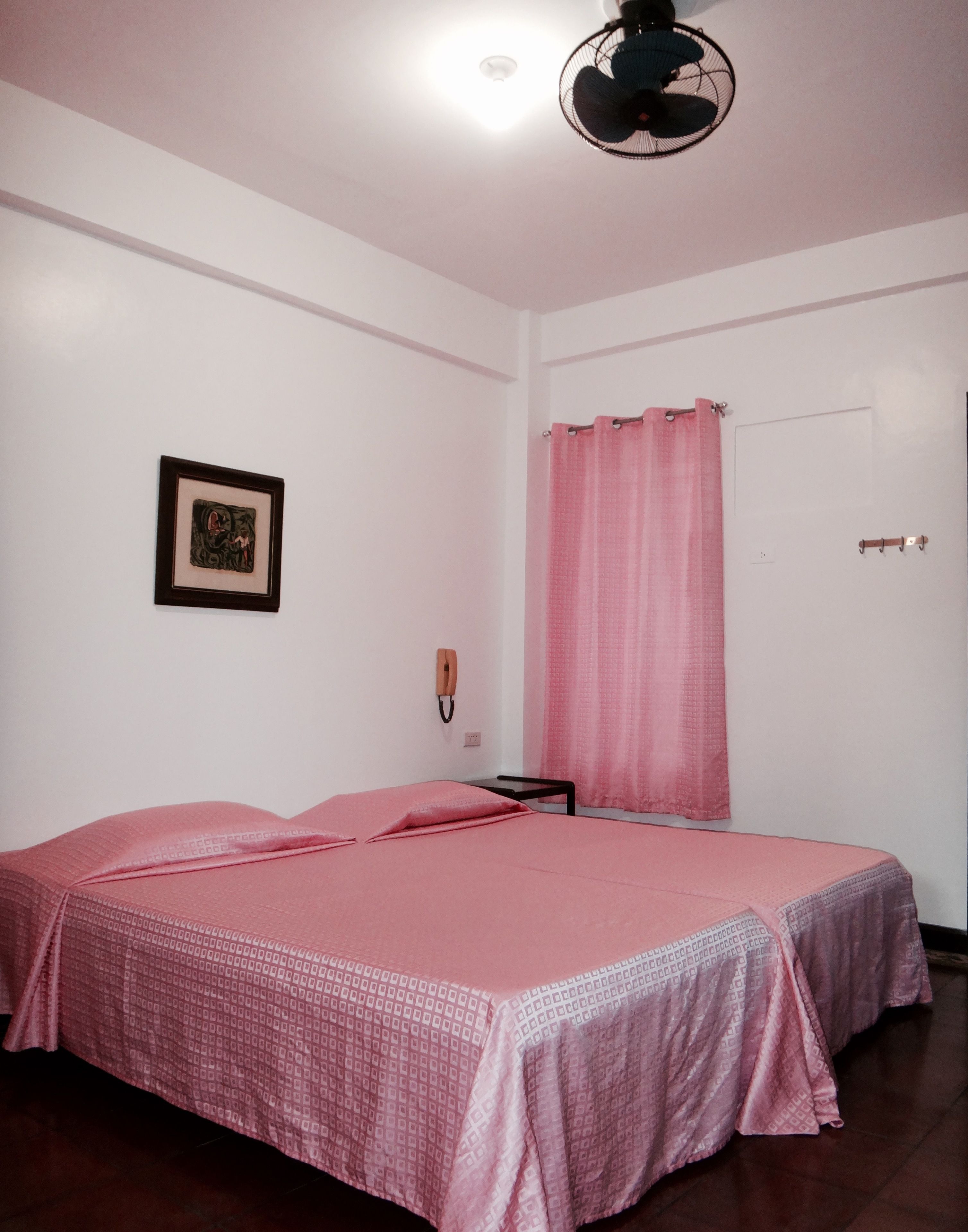 Quarto com duas camas, um ventilador de teto, um quadro na parede, um telefone e uma cortina rosa.