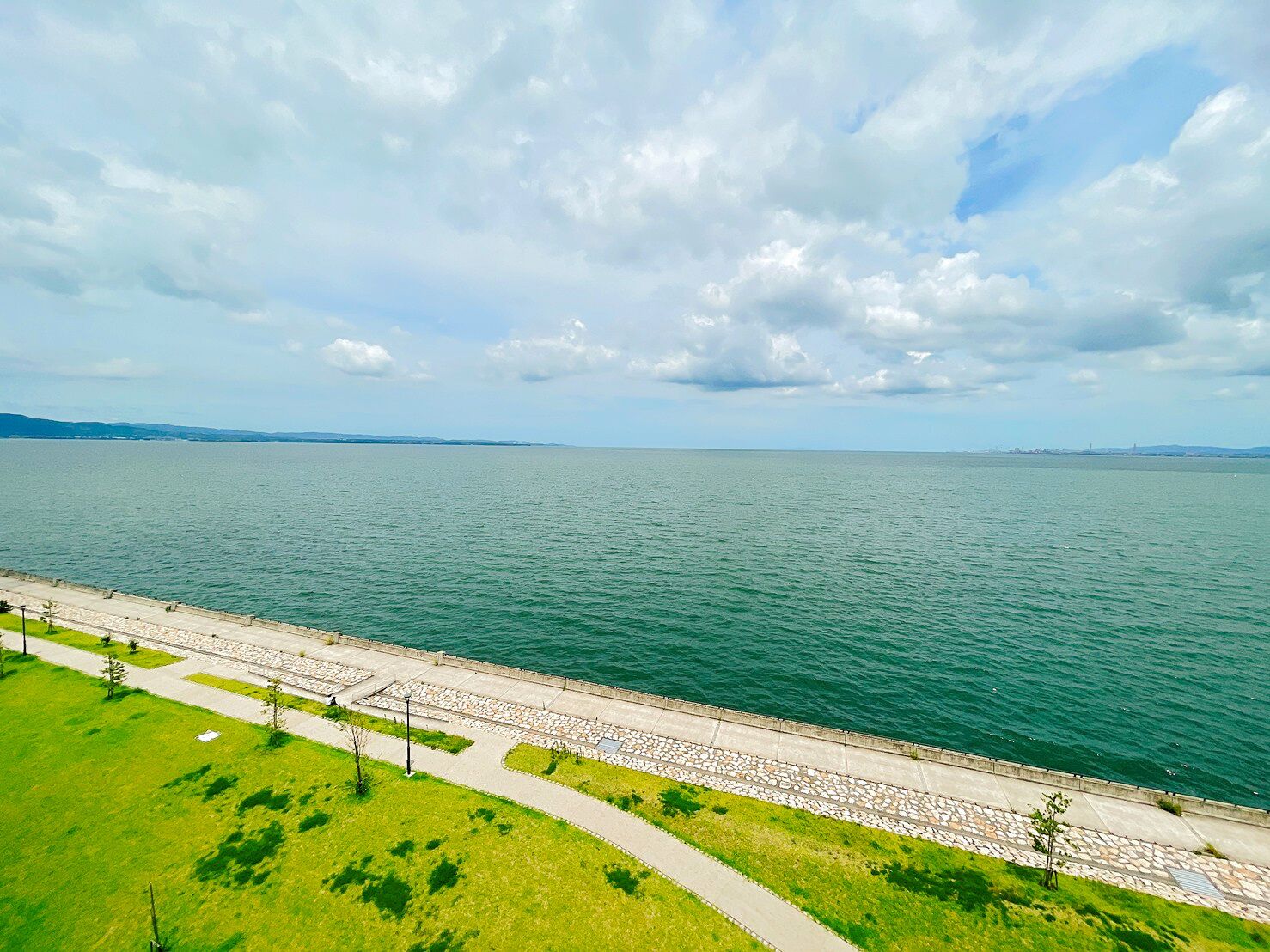Uma passarela costeira com limite de pedra, áreas ajardinadas e vista para o mar sob um céu parcialmente nublado.