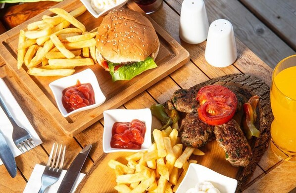Uma refeição com hambúrguer, batatas fritas e carne grelhada, acompanhada de uma fatia de tomate, servida sobre uma mesa de madeira com saleiro e pimenteiro, e um copo de suco de laranja.