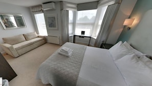 Quarto de hotel com cama, sofá, escrivaninha e janela com cortinas.