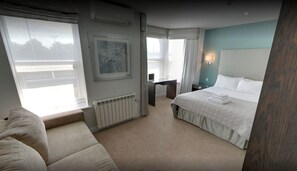 Quarto de hotel com uma cama grande, área de estar com sofá, escrivaninha e janela com cortinas.