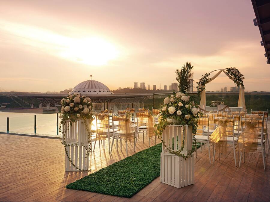 Decoração de casamento ao ar livre com cadeiras brancas e flores, com vista para uma paisagem urbana e um corpo d'água ao pôr do sol.