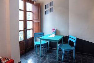 Um quarto pequeno com porta de madeira, janela, mesa com cadeiras e prateleira na parede.