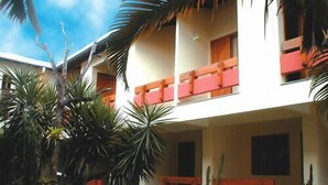 Um edifício com estrutura em dois andares, varandas com guarda-corpos vermelhos e um jardim com palmeiras e cactos.