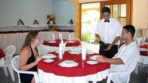 Uma área de refeições com mesas redondas cobertas por toalhas brancas e jogos americanos vermelhos, com cadeiras brancas.