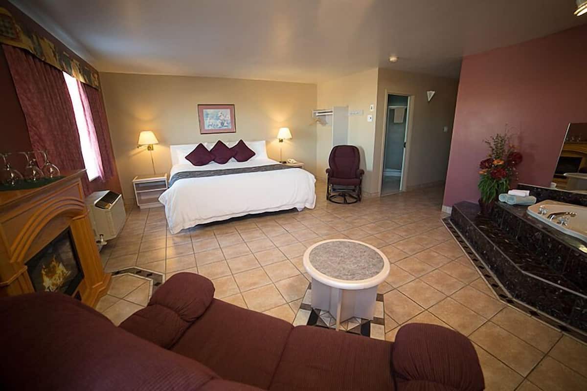 Quarto de hotel com cama, duas luminárias de cabeceira, uma cadeira, uma mesa redonda, lareira e banheiro com pia e espelho.