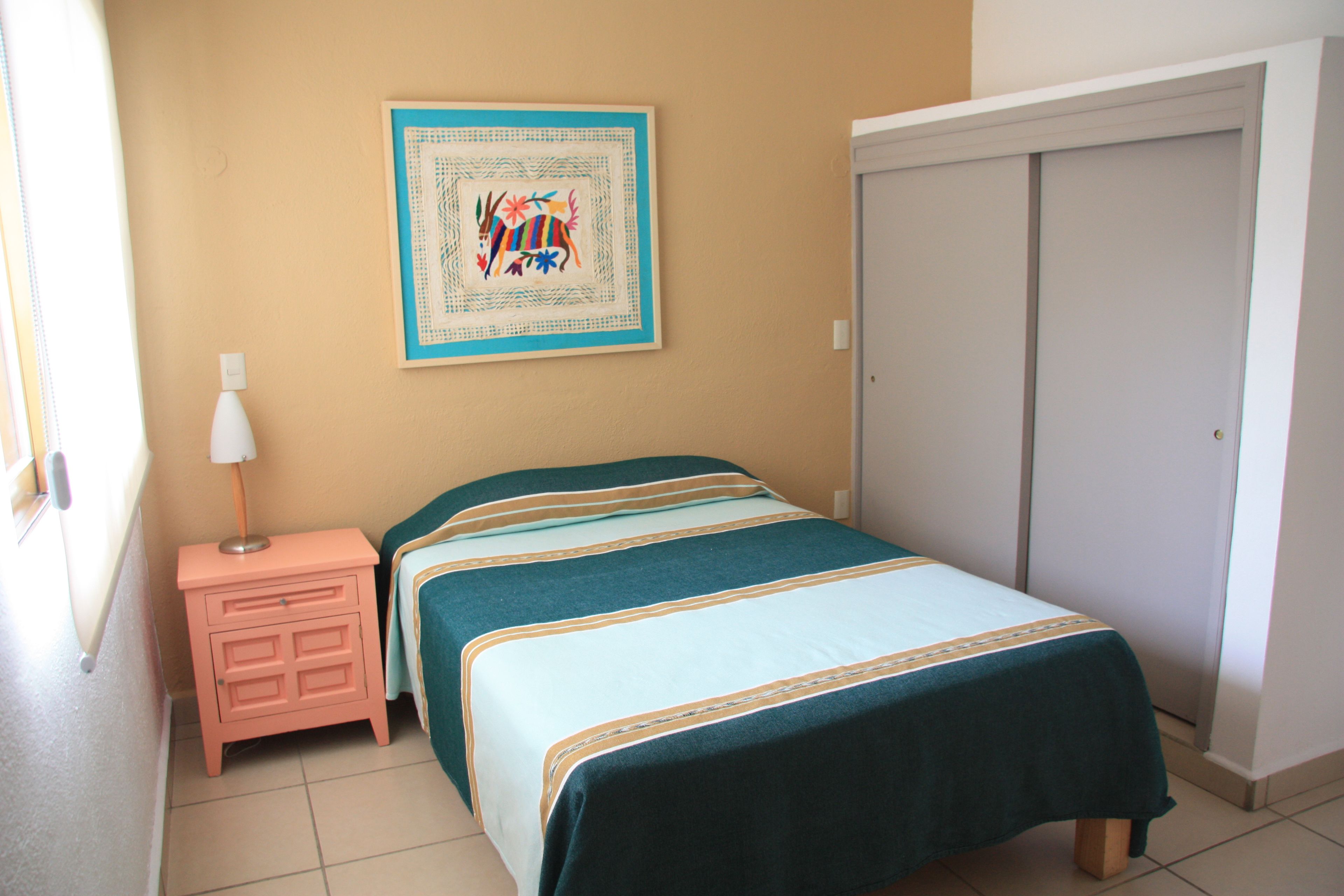 Quarto com cama, mesa de cabeceira, abajur, porta e uma obra de arte na parede.