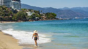 Uma praia com uma pessoa caminhando em direção à água, edifícios ao fundo e montanhas à distância.