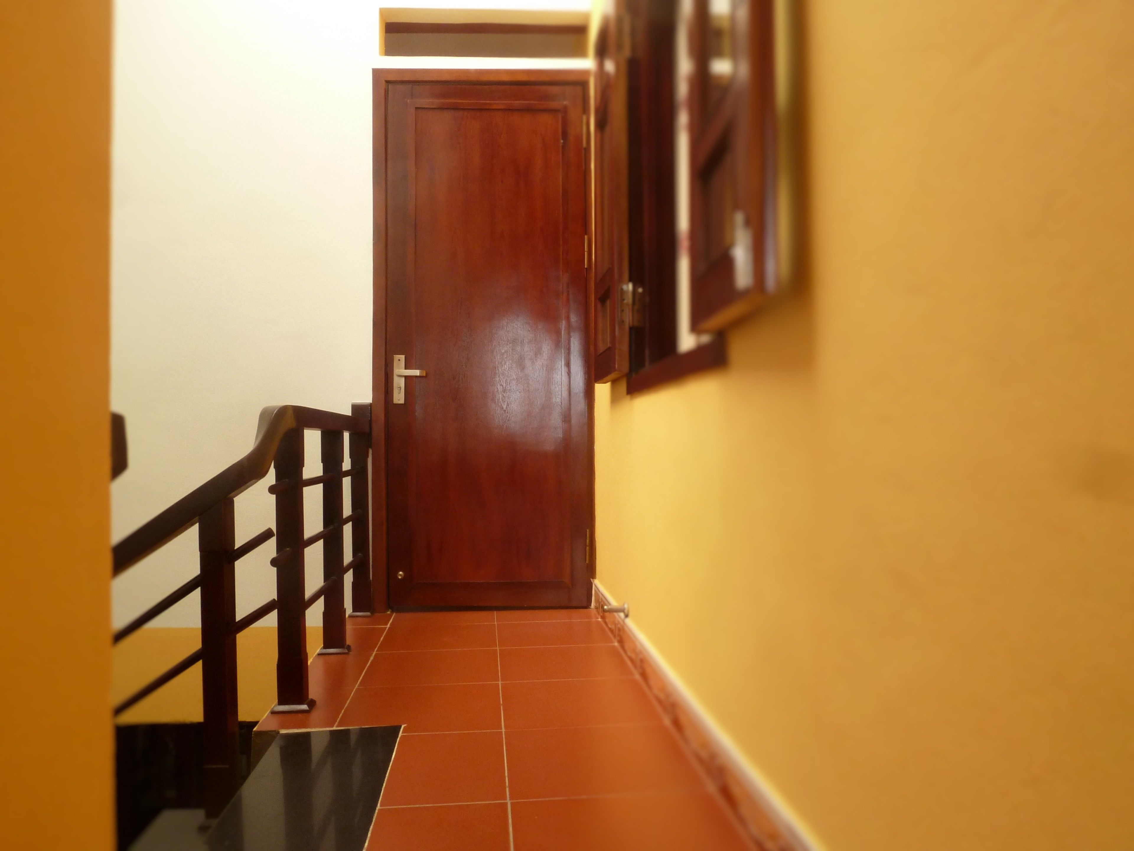 Um corredor com porta de madeira, tapete preto na entrada e chão ladrilhado.