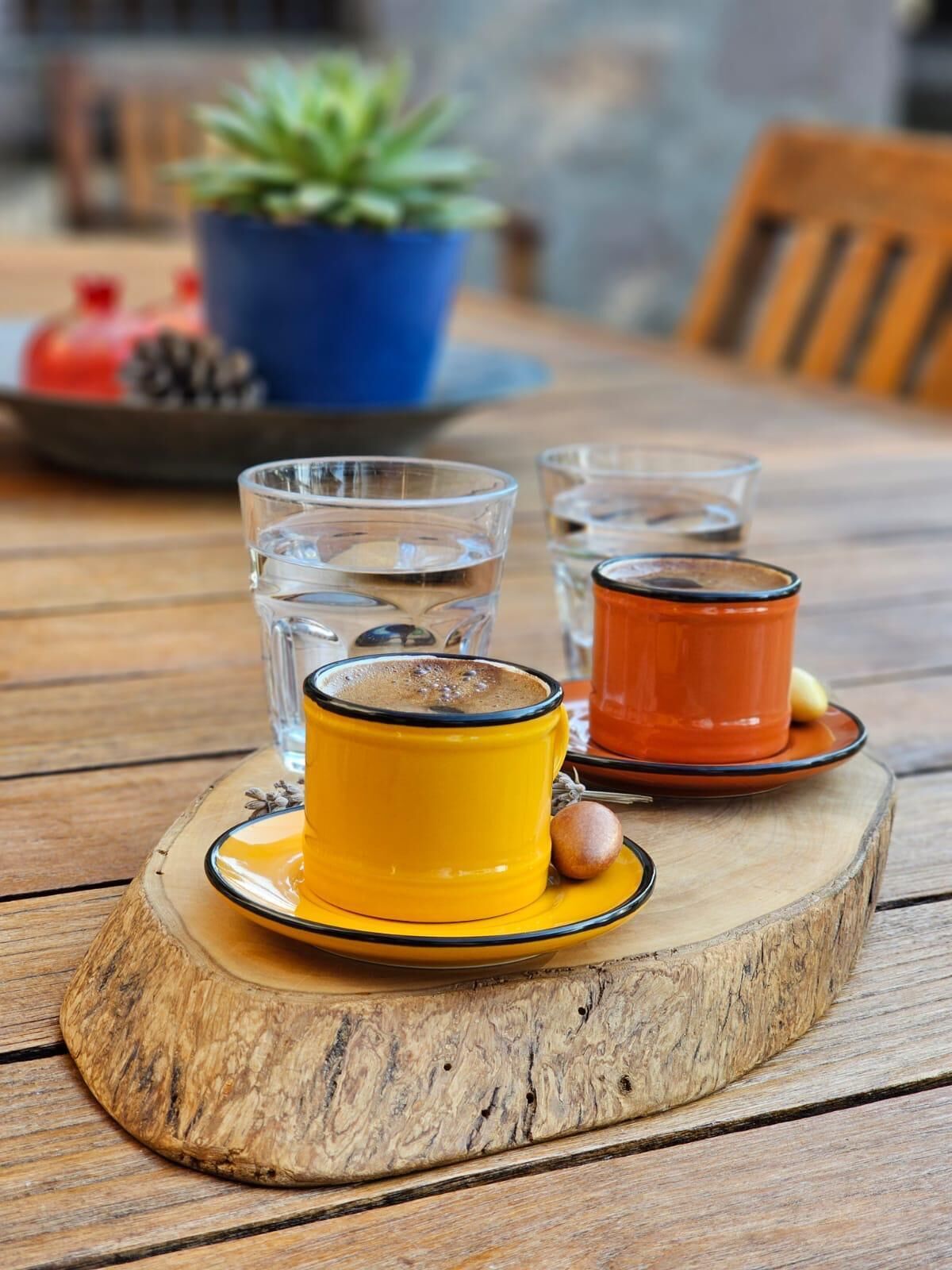 Uma mesa de madeira com uma caneca amarela e um copo de vidro, um vaso azul com planta e uma cadeira de madeira ao fundo.