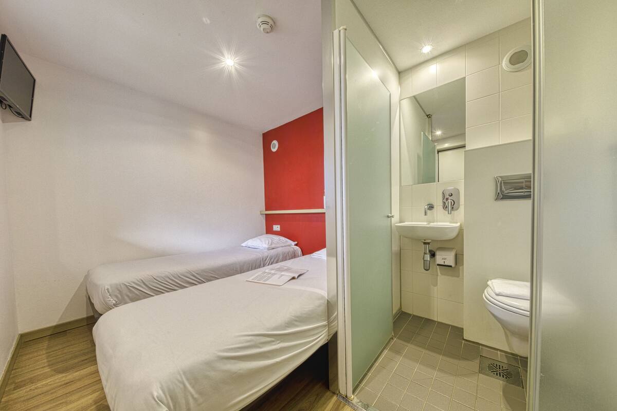 Quarto de hotel com duas camas de solteiro, uma parede de destaque vermelha, um banheiro com pia e vaso sanitário, e um espelho.