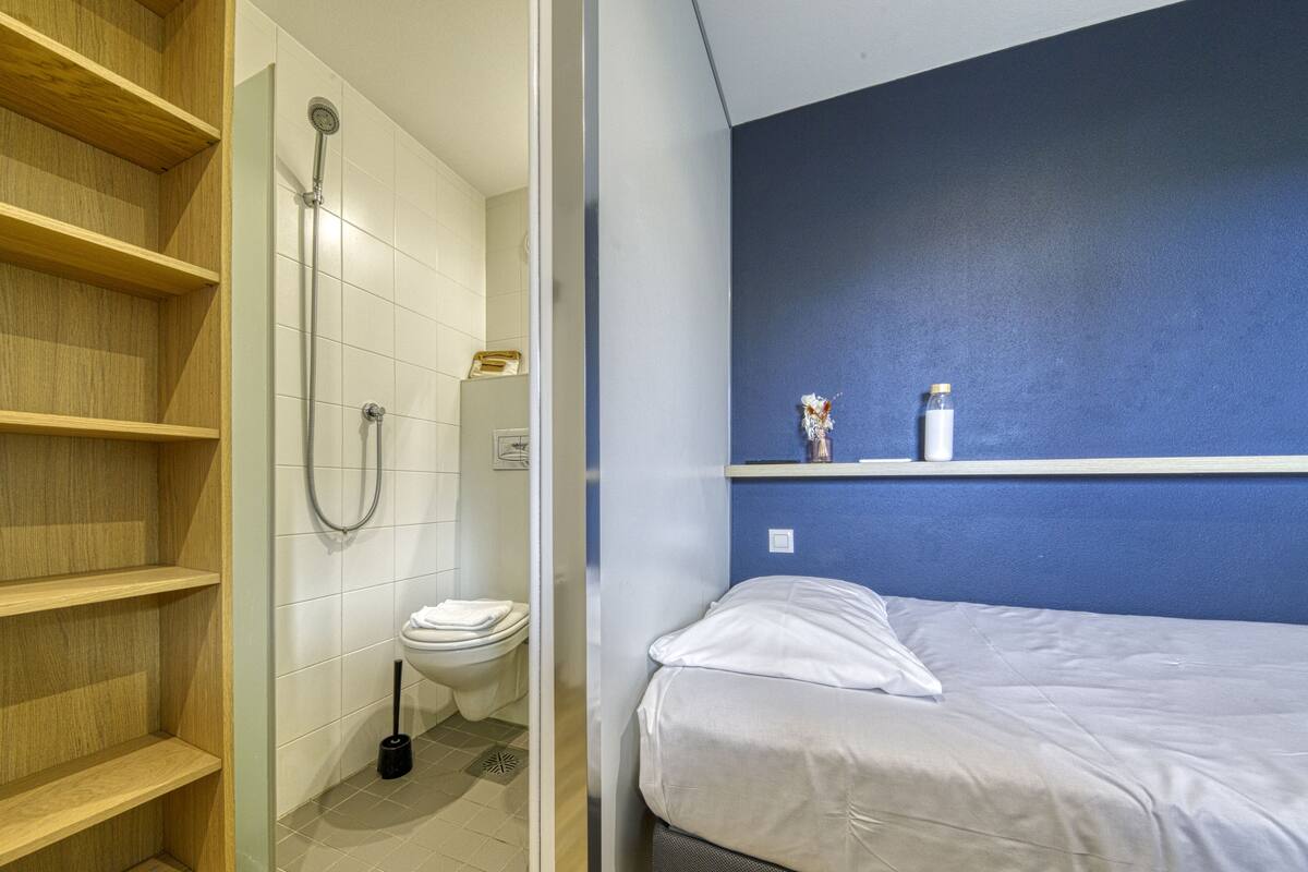 Casa de banho com chuveiro, sanita e cama, parede azul e estantes.