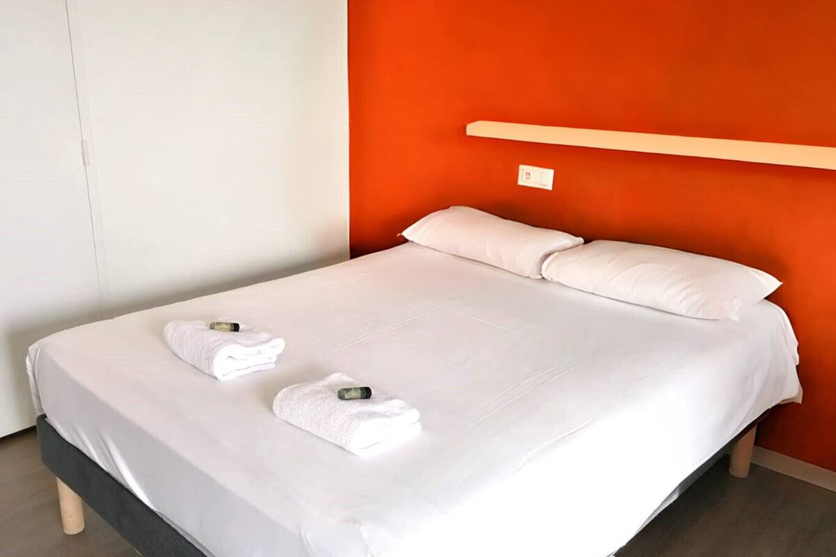Cama bem arrumada com lençóis brancos e duas toalhas, uma parede branca e uma parede de destaque laranja.