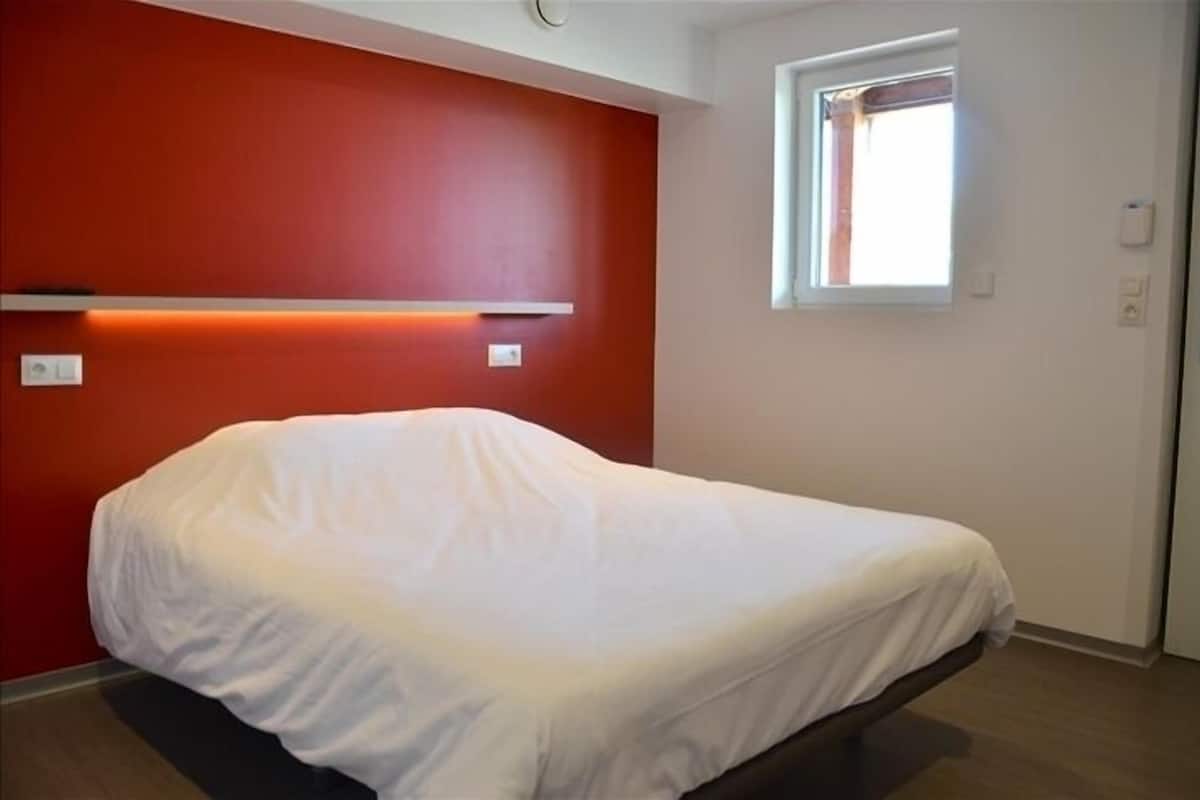 Uma cama de solteiro num quarto com uma parede em vermelho e uma janela.