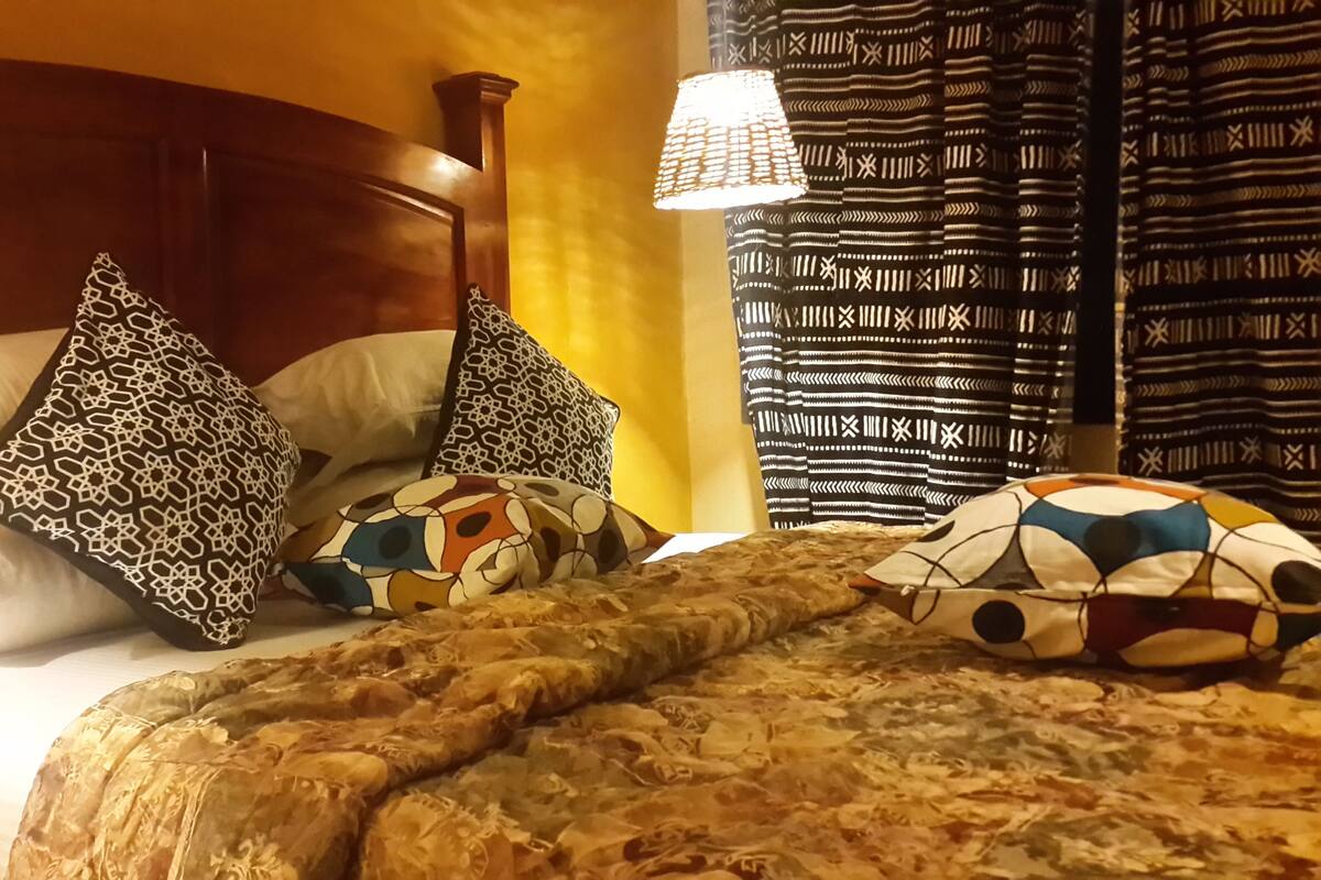 Cama com almofadas estampadas, cabeceira de madeira, luminária e cortinas padronizadas.