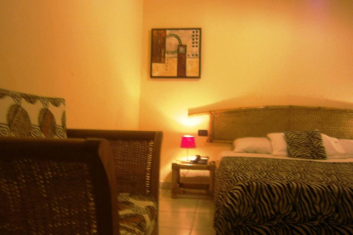 Quarto de hotel com cama, uma cadeira, mesa de cabeceira e um quadro na parede.