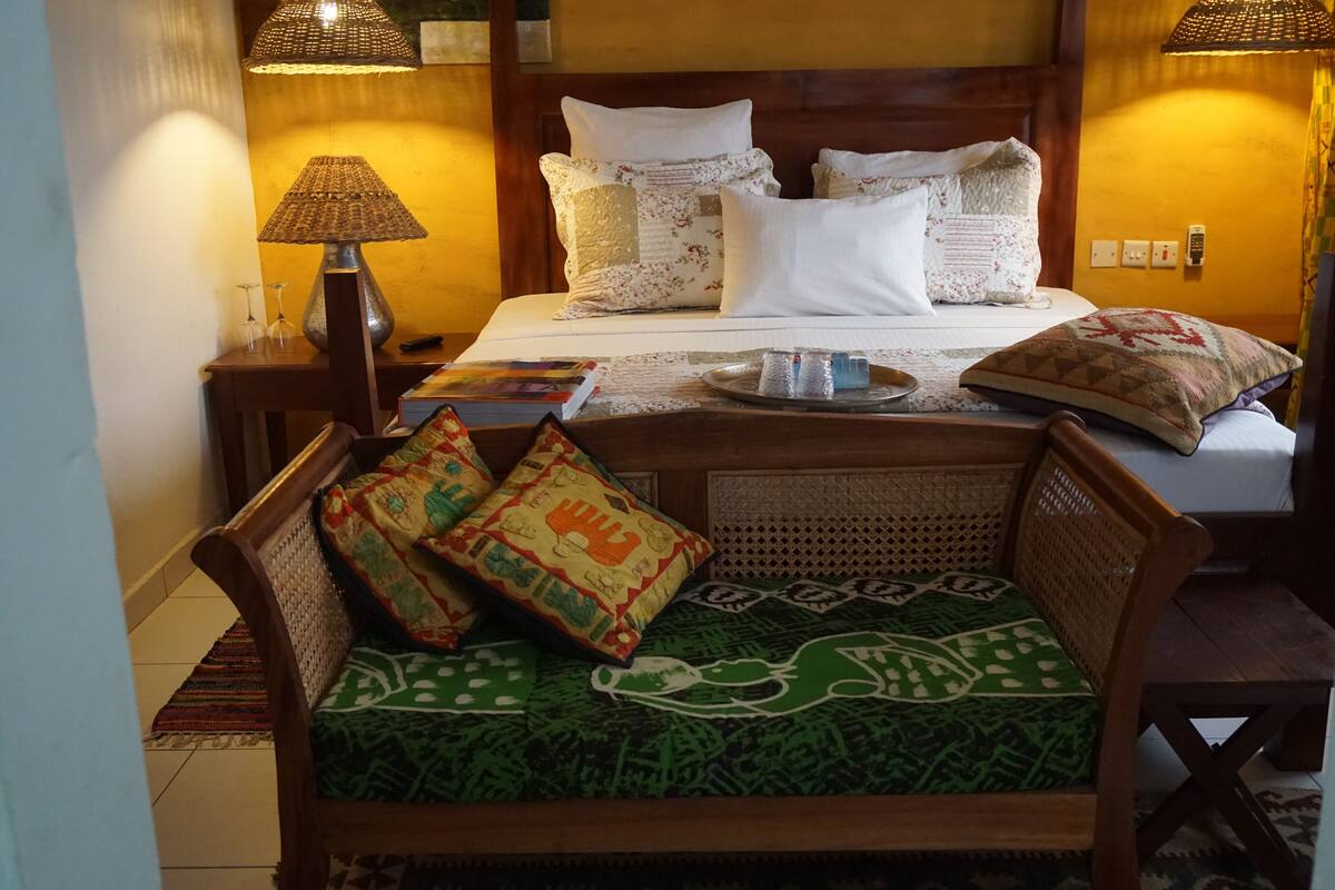 Quarto de hotel com cama, um banquinho de madeira com almofadas, luminária e bandeja com garrafa e copos.