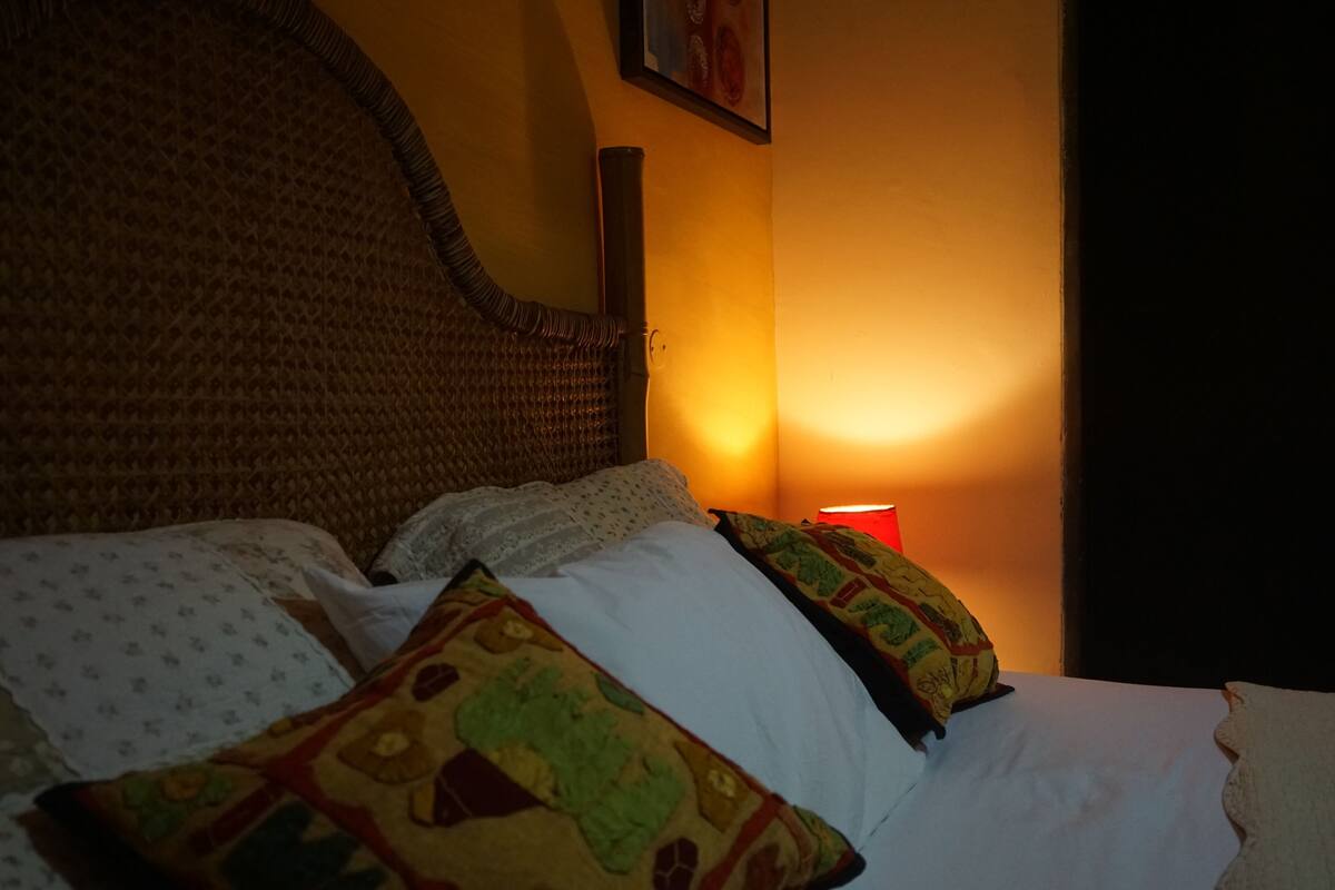 Uma cama com almofadas estampadas e cabeceira, um abajur e um quadro na parede.