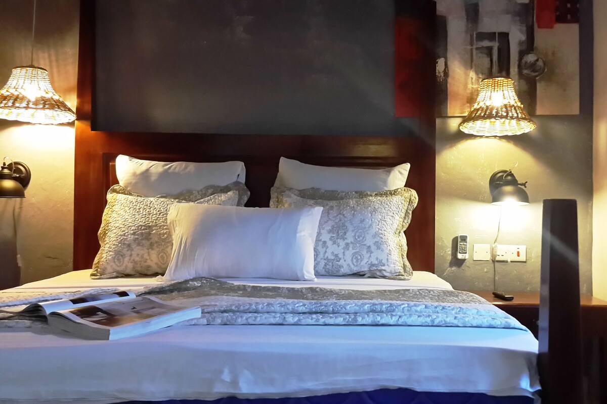 Cama bem arrumada com colcha branca e azul, várias almofadas e duas luminárias fixadas na parede, iluminando o ambiente.