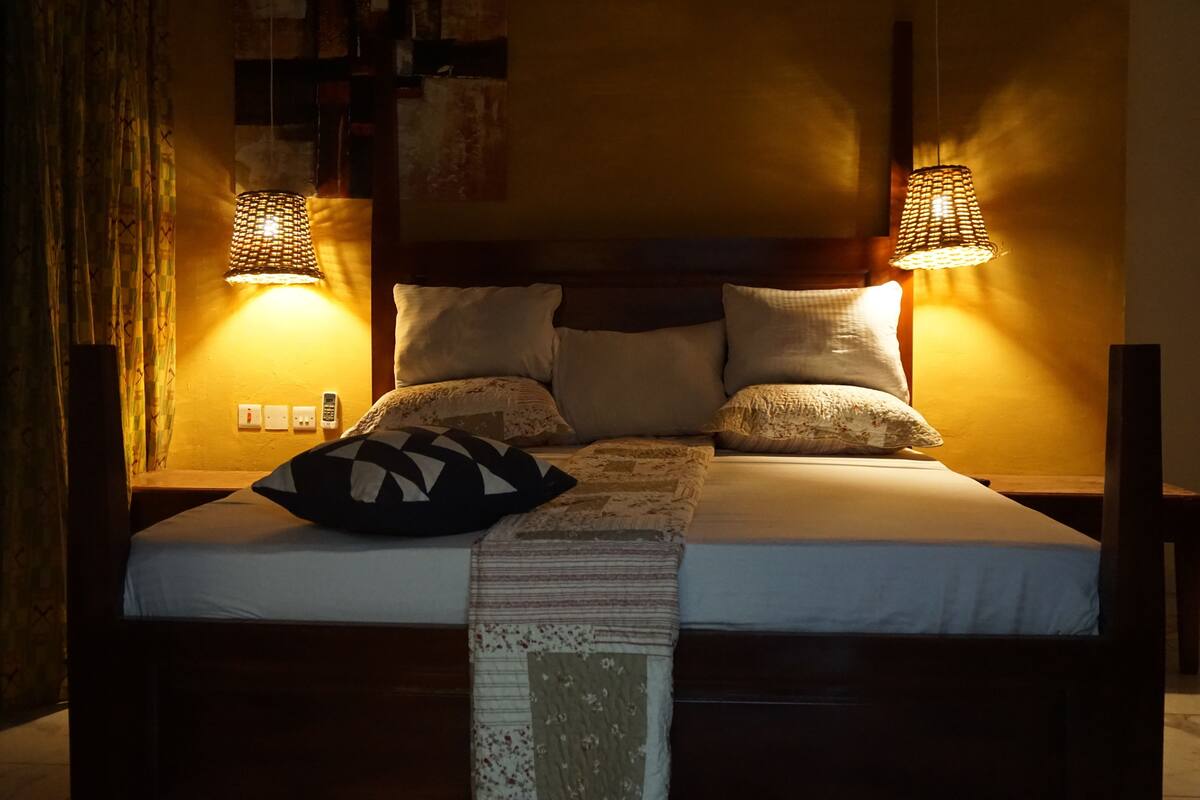 Cama bem arrumada com manta e almofadas estampadas, duas luminárias na parede e uma tomada.