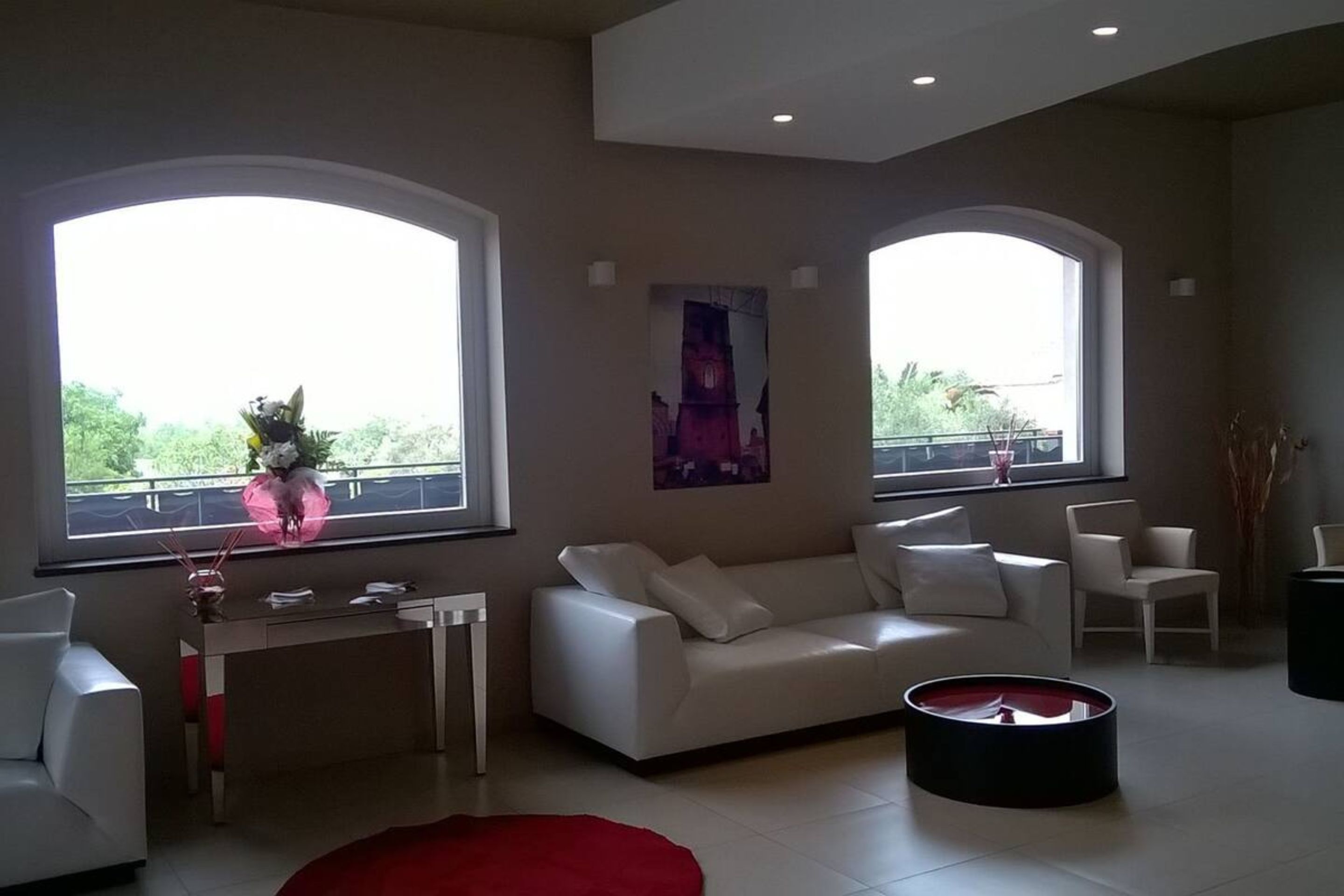 Uma sala de estar moderna com um sofá branco, um banquinho vermelho e uma mesa de centro redonda. Há duas janelas com arcos, e uma pintura é visível na parede.