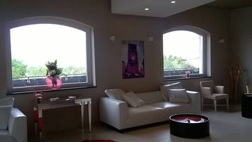 Uma sala de estar moderna com um sofá branco, um banquinho vermelho e uma mesa de centro redonda. Há duas janelas com arcos, e uma pintura é visível na parede.