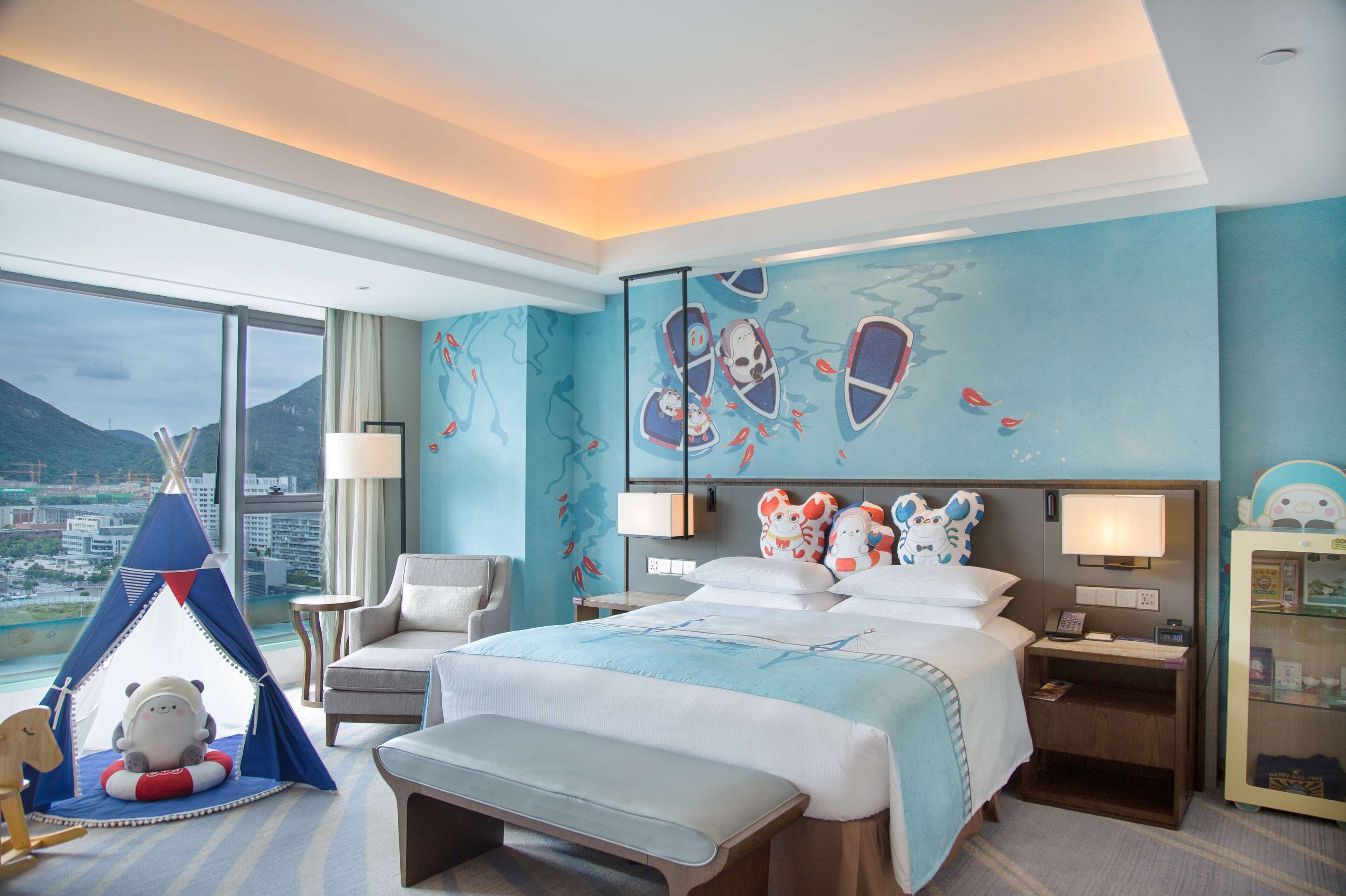 Quarto de hotel com uma cama grande, uma escrivaninha, uma cadeira, uma mesinha, um abajur, um guarda-roupa e vista para a cidade.