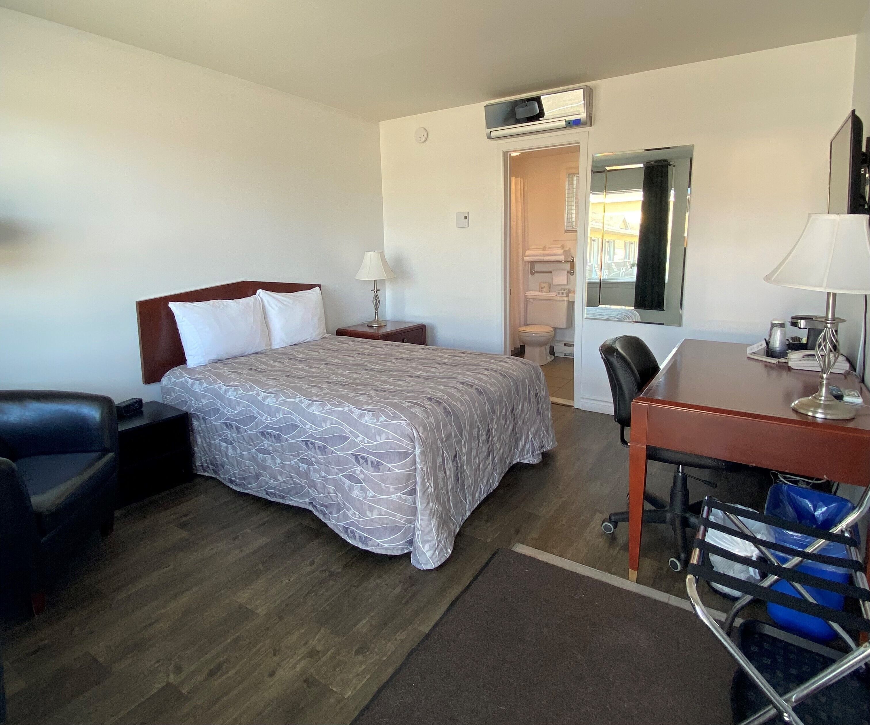Quarto de hotel com cama, escrivaninha, cadeira, mesa de cabeceira, luminária, espelho e banheiro visível por uma porta aberta.