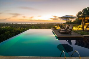 Uma piscina infinita com deck de madeira e espreguiçadeiras, com vista para uma paisagem arborizada.
