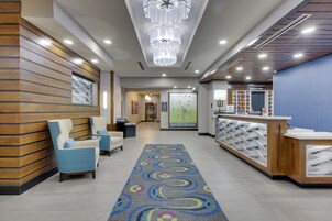 Uma área de recepção de hotel com design moderno, apresentando um balcão de recepção, área de estar com poltronas, um tapete estampado e uma decoração de parede com tema esportivo.