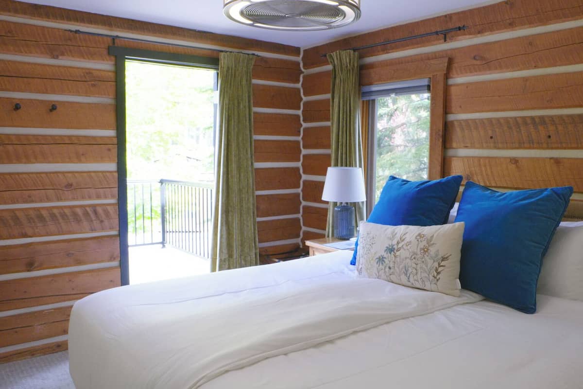 Quarto de cabana de toras com uma cama grande, uma mesinha de cabeceira com abajur e uma janela com cortinas.