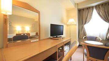 Quarto de hotel com escrivaninha de madeira, televisão, cadeira, mesa pequena e janela com cortinas.