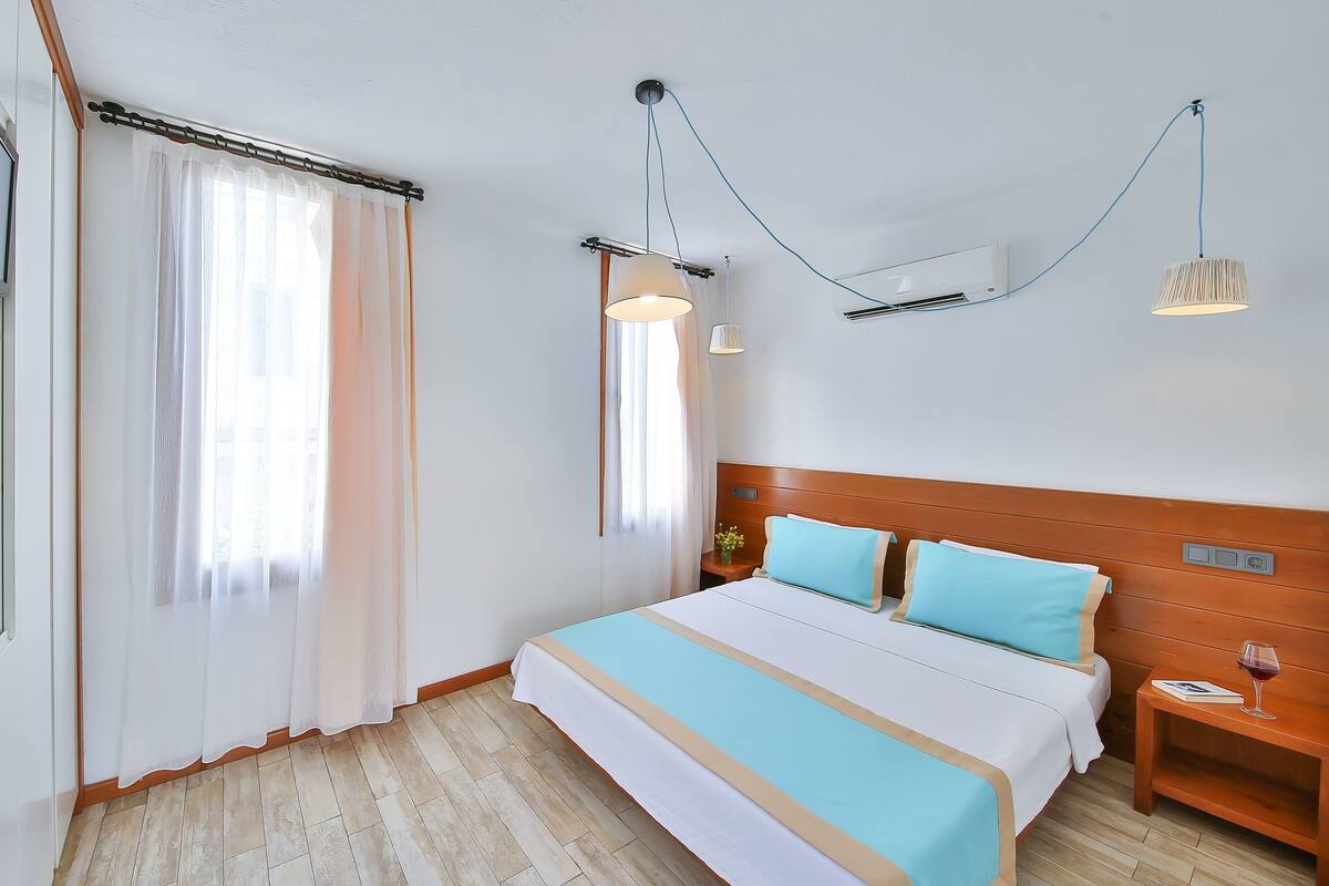 Quarto de hotel com cama, cabeceira de madeira, mesa de cabeceira e luminária pendente.