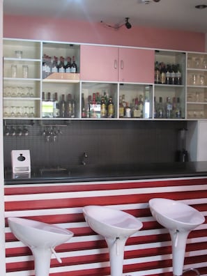 Uma área de bar com prateleiras de garrafas e copos, um revestimento de azulejos e três banquetas brancas.
