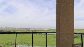 Varanda com uma poltrona estofada vermelha, uma cadeira de vime e vista para uma paisagem verde e edifícios ao longe.