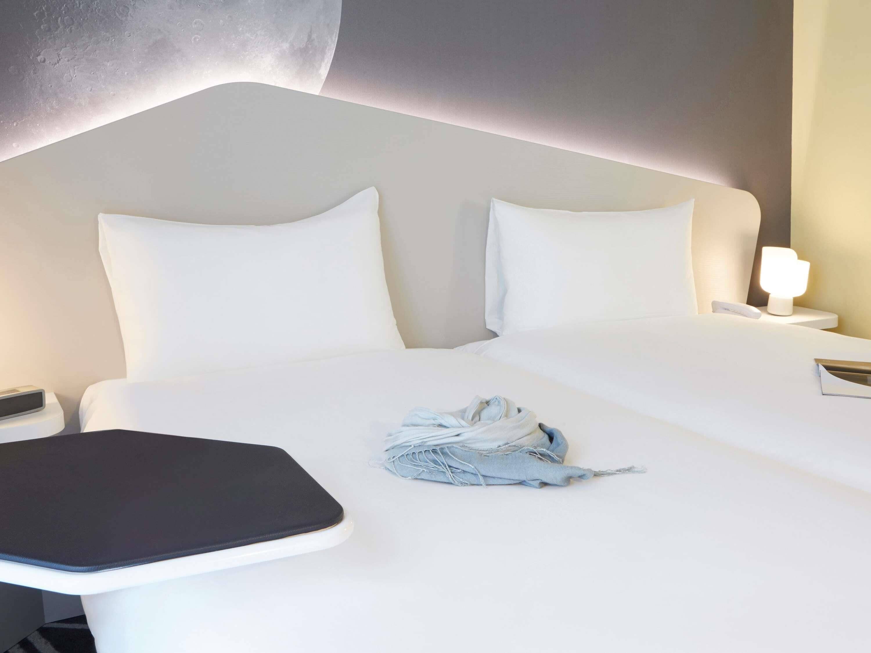 Quarto de hotel com cama, mesa de cabeceira e criado-mudo com luminária.