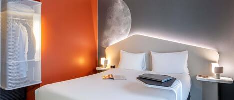 Quarto de hotel moderno com uma cama grande, mesa de cabeceira com luminária e televisão na parede.