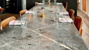 Uma longa mesa de jantar com cadeiras, um lustre e uma parede de tijolos ao fundo.
