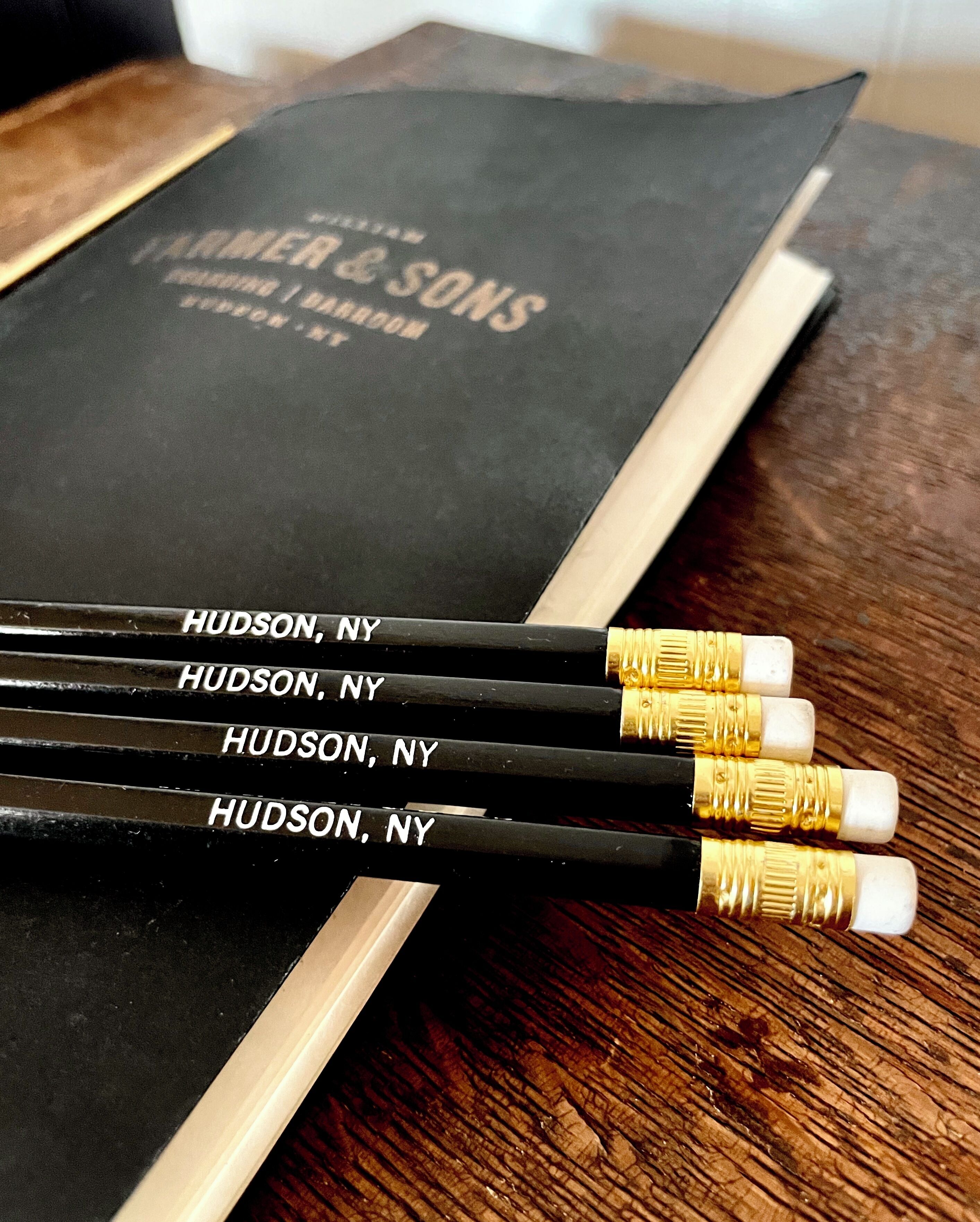 Um livro com o título 'JAMES & SONS' e quatro lápis com a inscrição 'HUDSON, NY' sobre uma superfície de madeira.