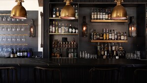 Uma área de bar com balcão de madeira escura, diversas prateleiras com garrafas de bebidas e taças de vinho penduradas.